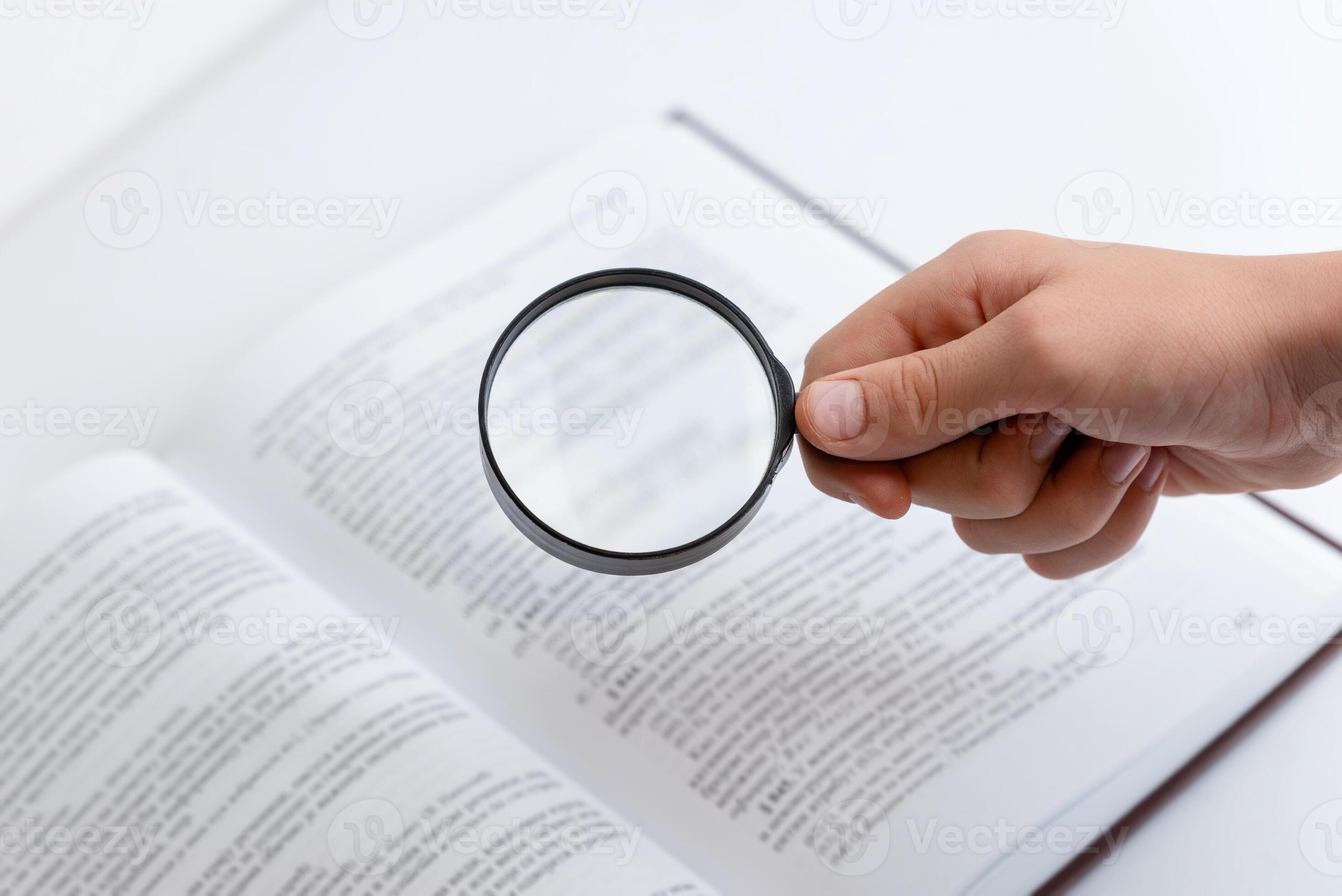 Hand holding magnifying glass searching through open book with dense text, symbolizing research ...