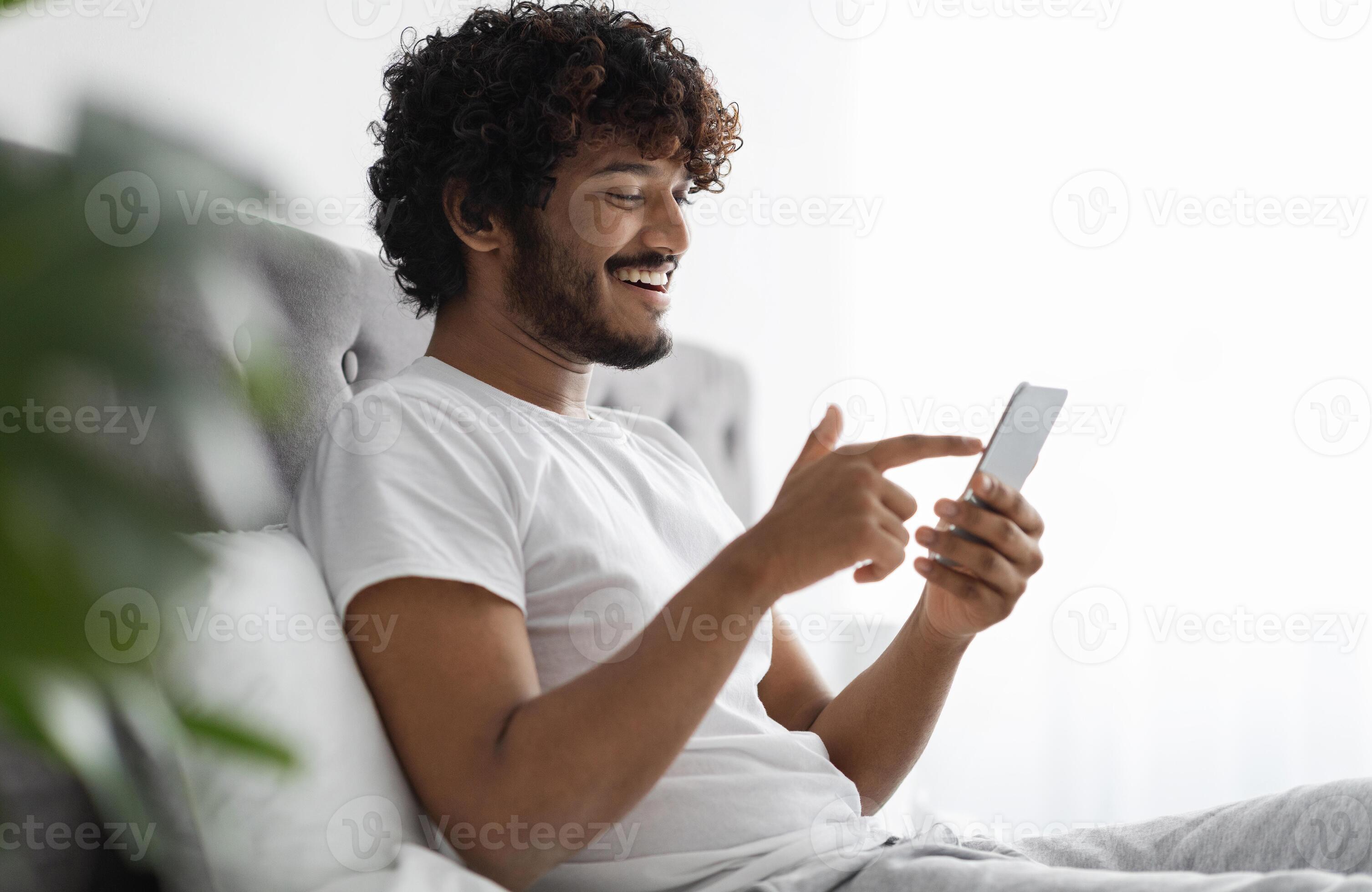 Positive curly bearded millennial dark-skinned guy scrolling in bed, using smartphone, touching ...