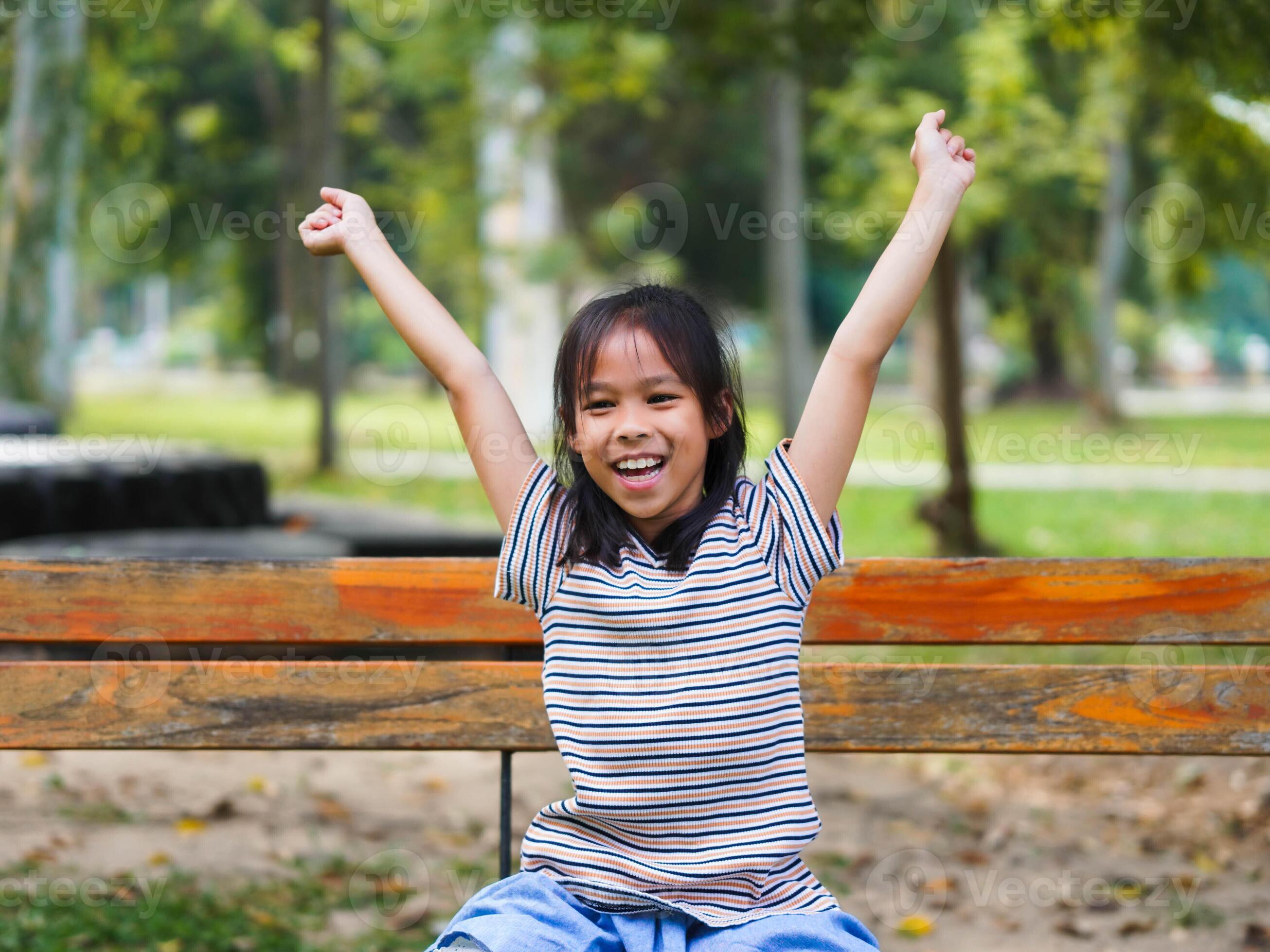 A happy little girl is sitting on a park bench. Smiling little girl with arms outstretched ...