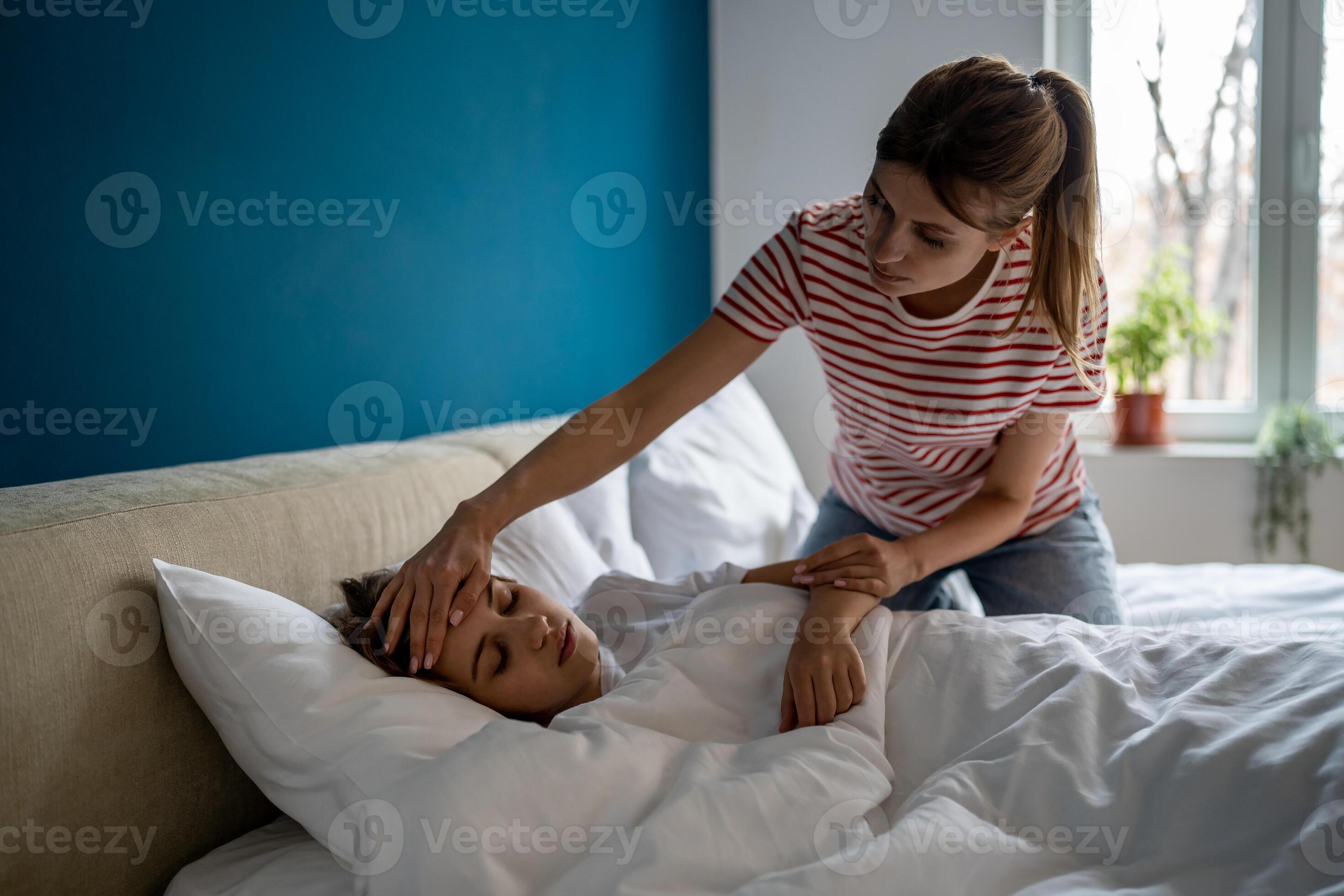 Sick teenage girl lying in bed, having chills and fever, mom checking temperature touching ...