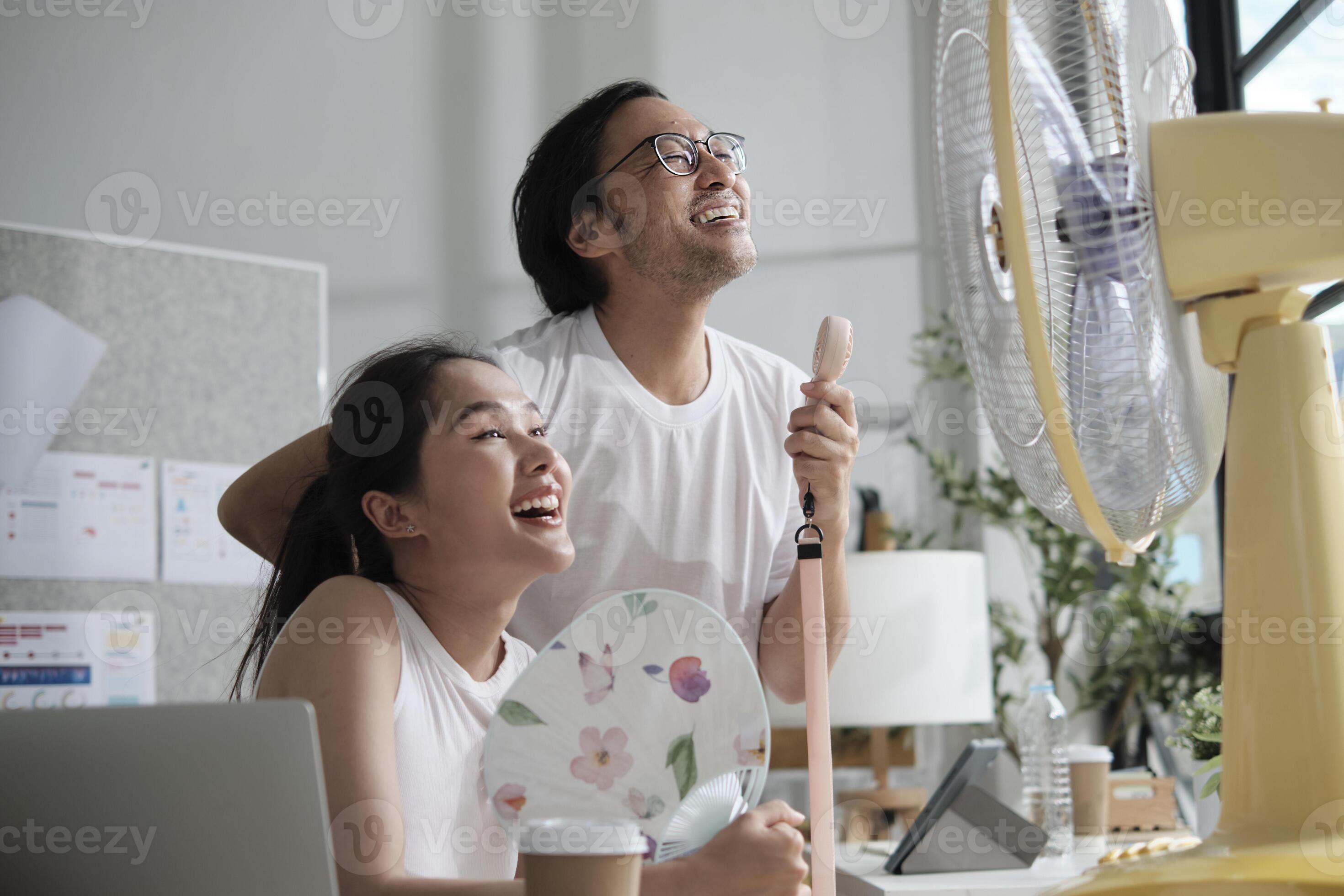 Two young Asian SME coworkers are fun blowing wind with electric fan, working in hot ...
