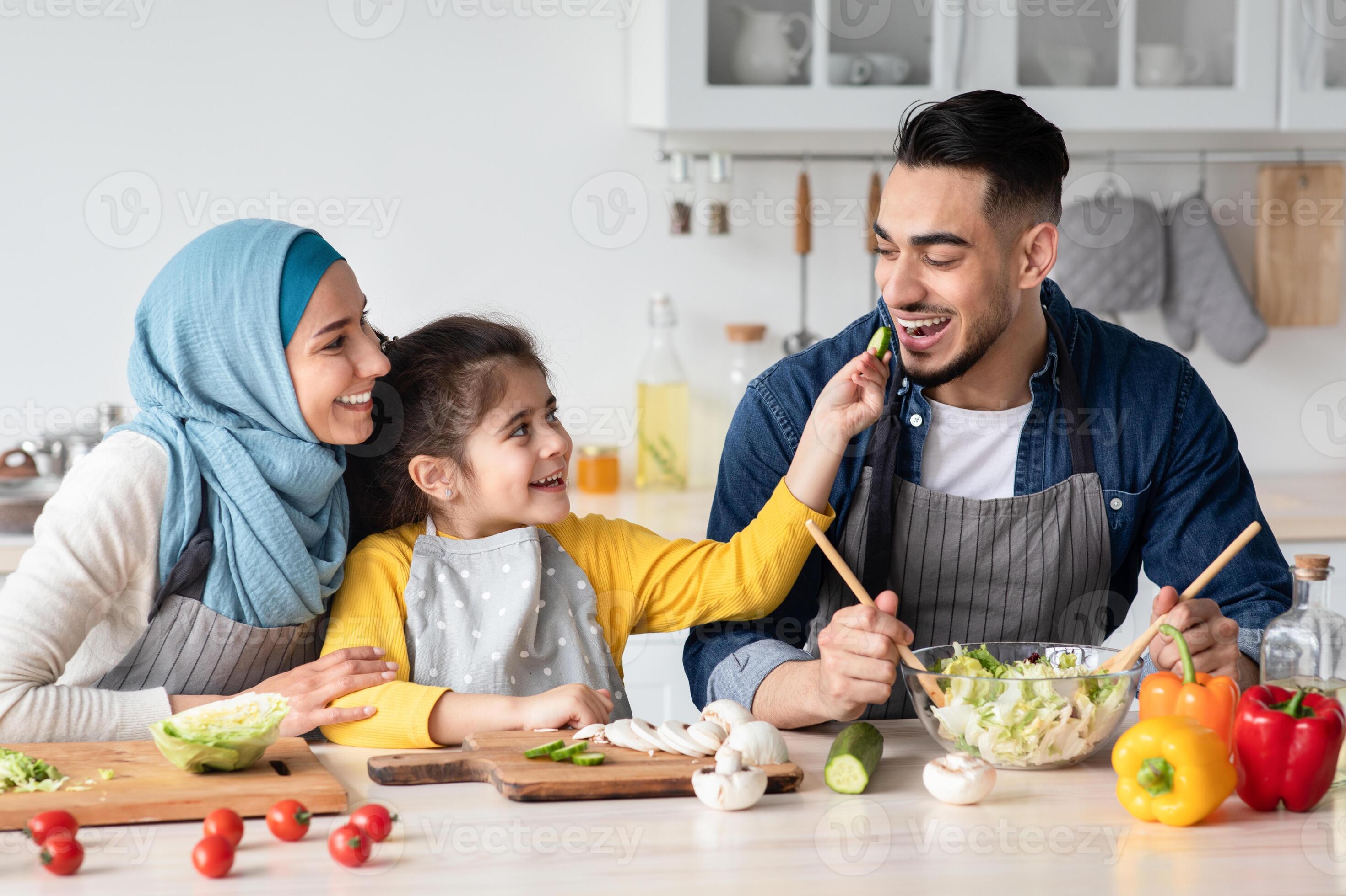 Taste It. Portrait of cute little arab girl feeding her daddy with cucumber slice, happy middle ...