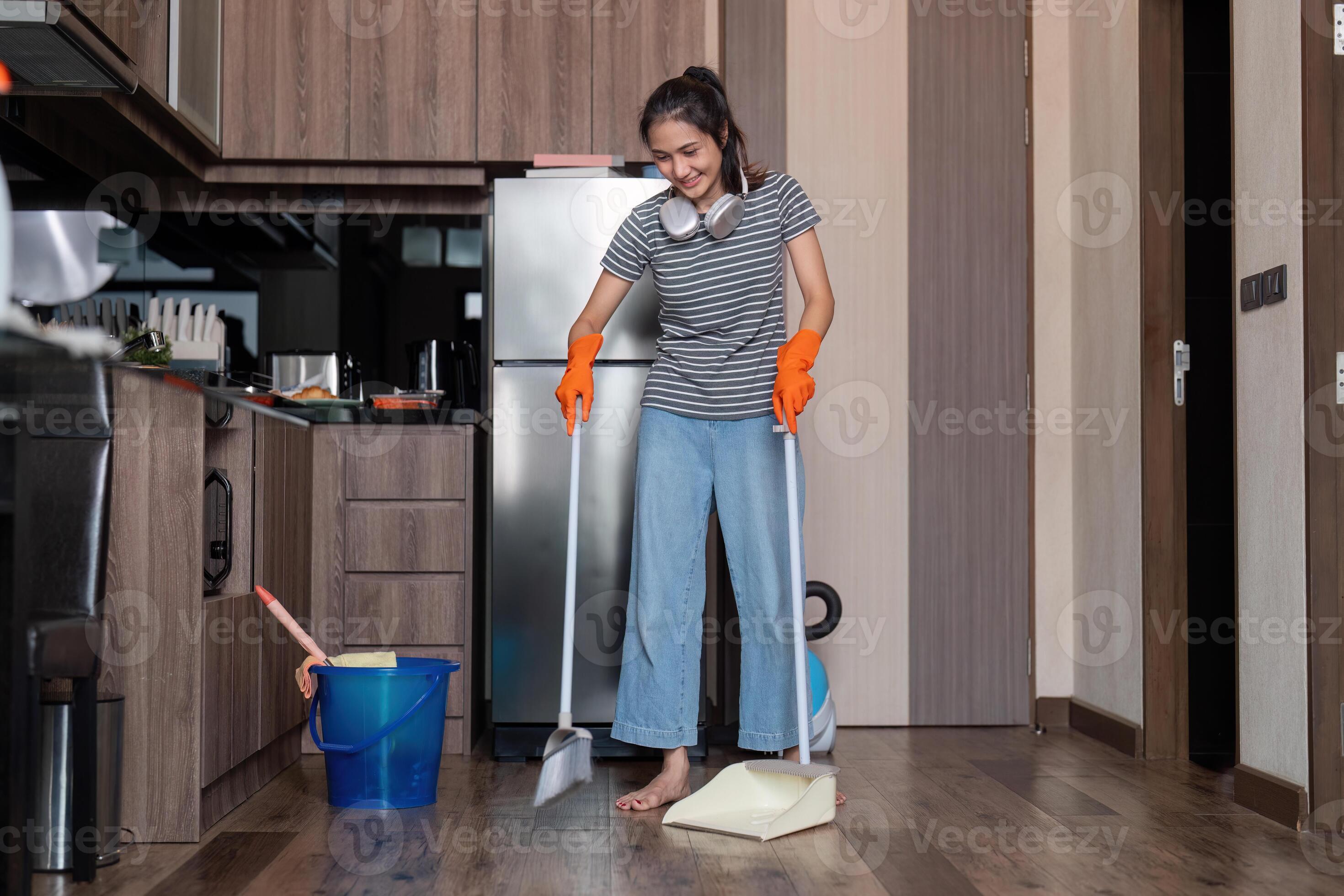 Young woman cleaning her kitchen floor, wearing orange gloves and smiling while using broom and ...
