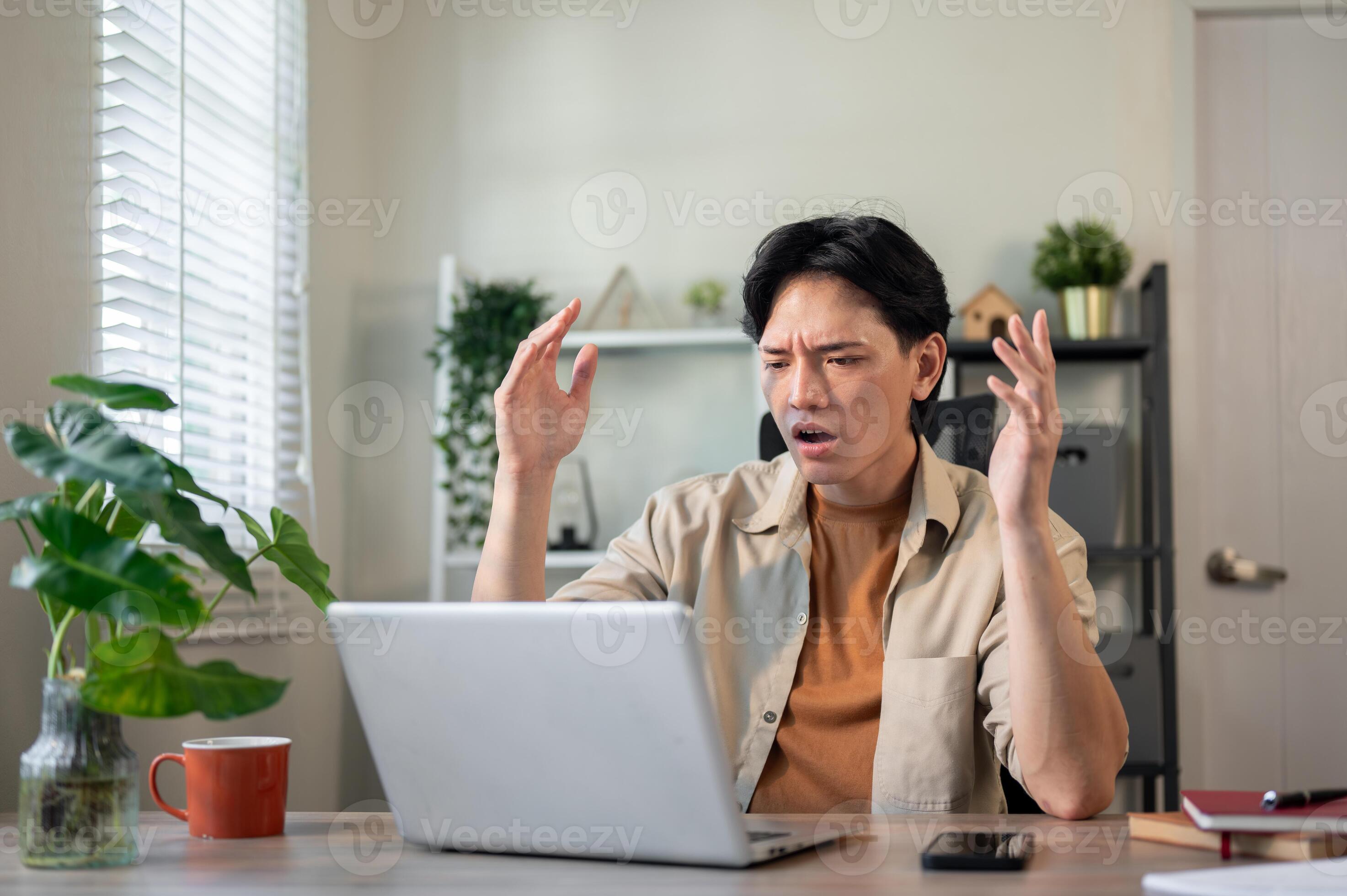 Angry Asian businessman sits at his desk, looking at his laptop screen ...