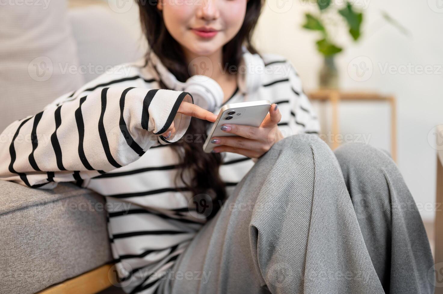 A close-up of an Asian woman is scrolling through her smartphone while relaxing in her living ...
