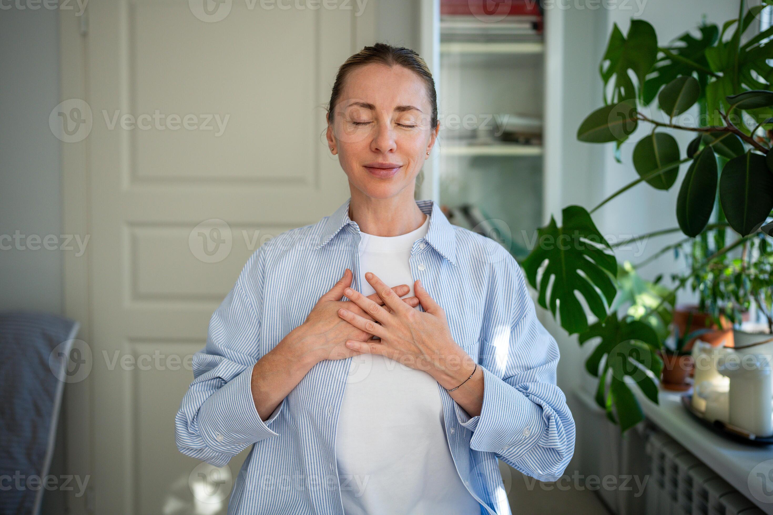 Peaceful woman standing with closed eyes, folding one's arms on chest doing breathing exercises ...
