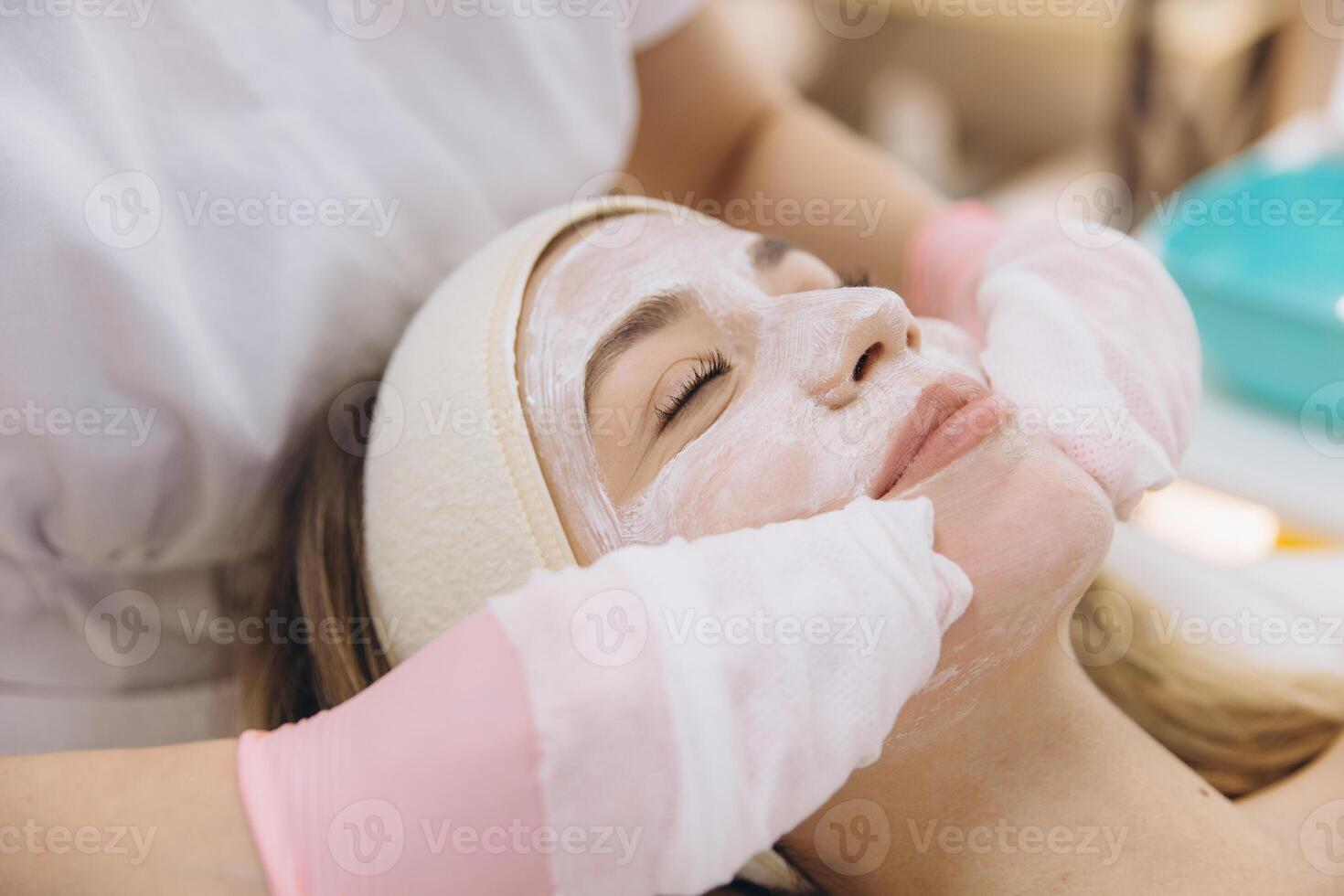 Beautician wearing gloves applying a white facial mask on the face of a young woman lying on a ...
