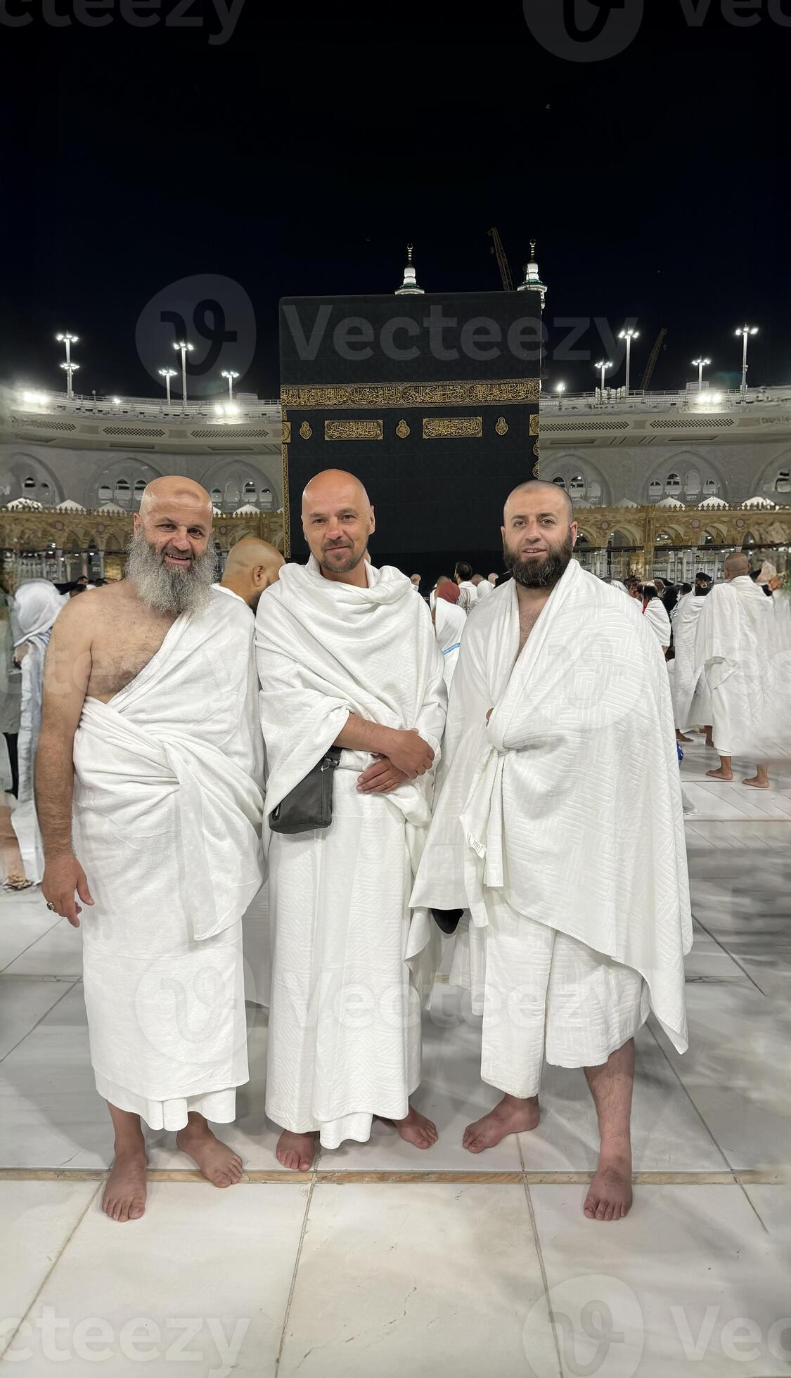 Three Muslim men in Ihram clothing standing in front of the Kaaba during Hajj or Umrah ...