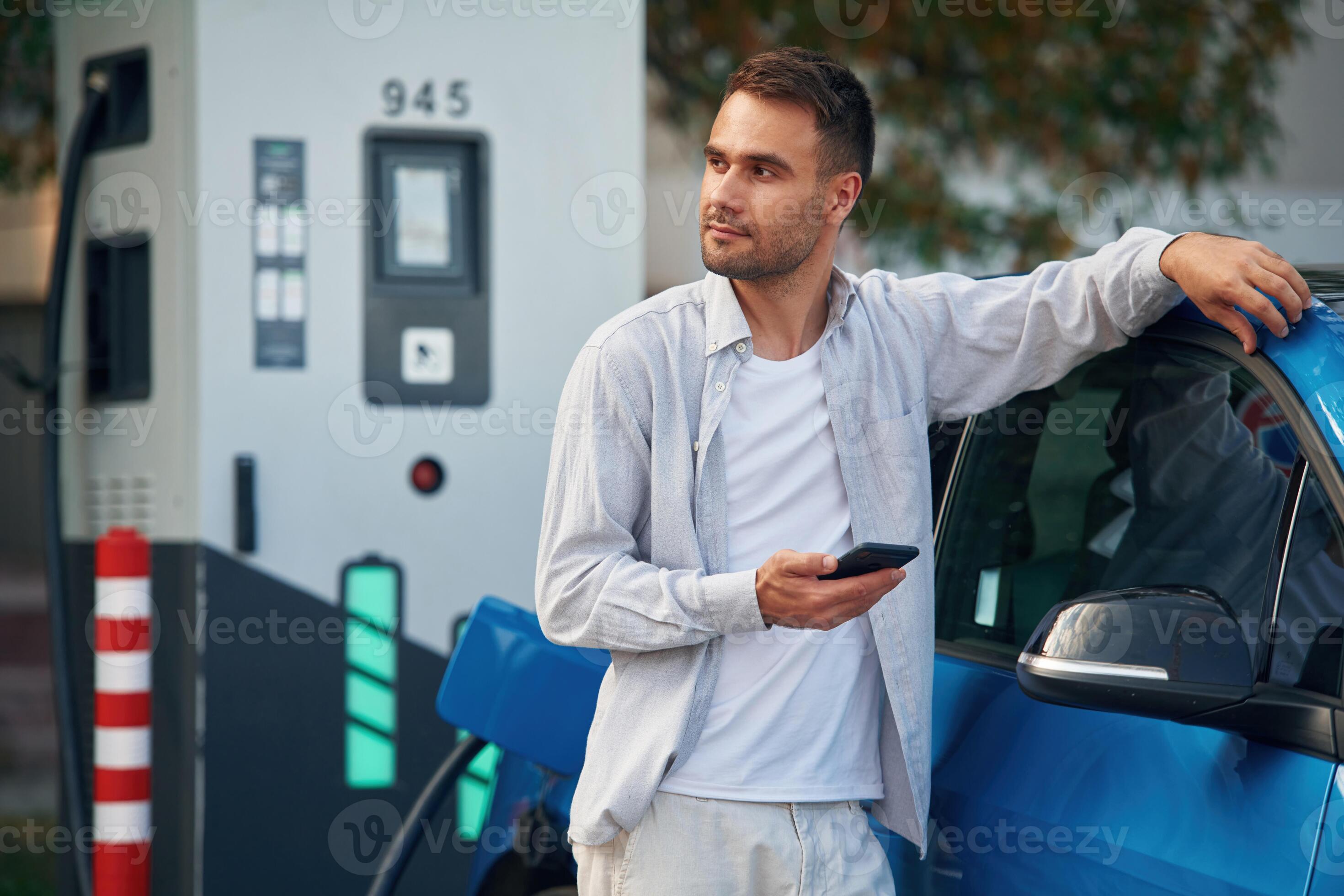 Standing and, waiting for process finished. Man with blue electric car on the charge station ...