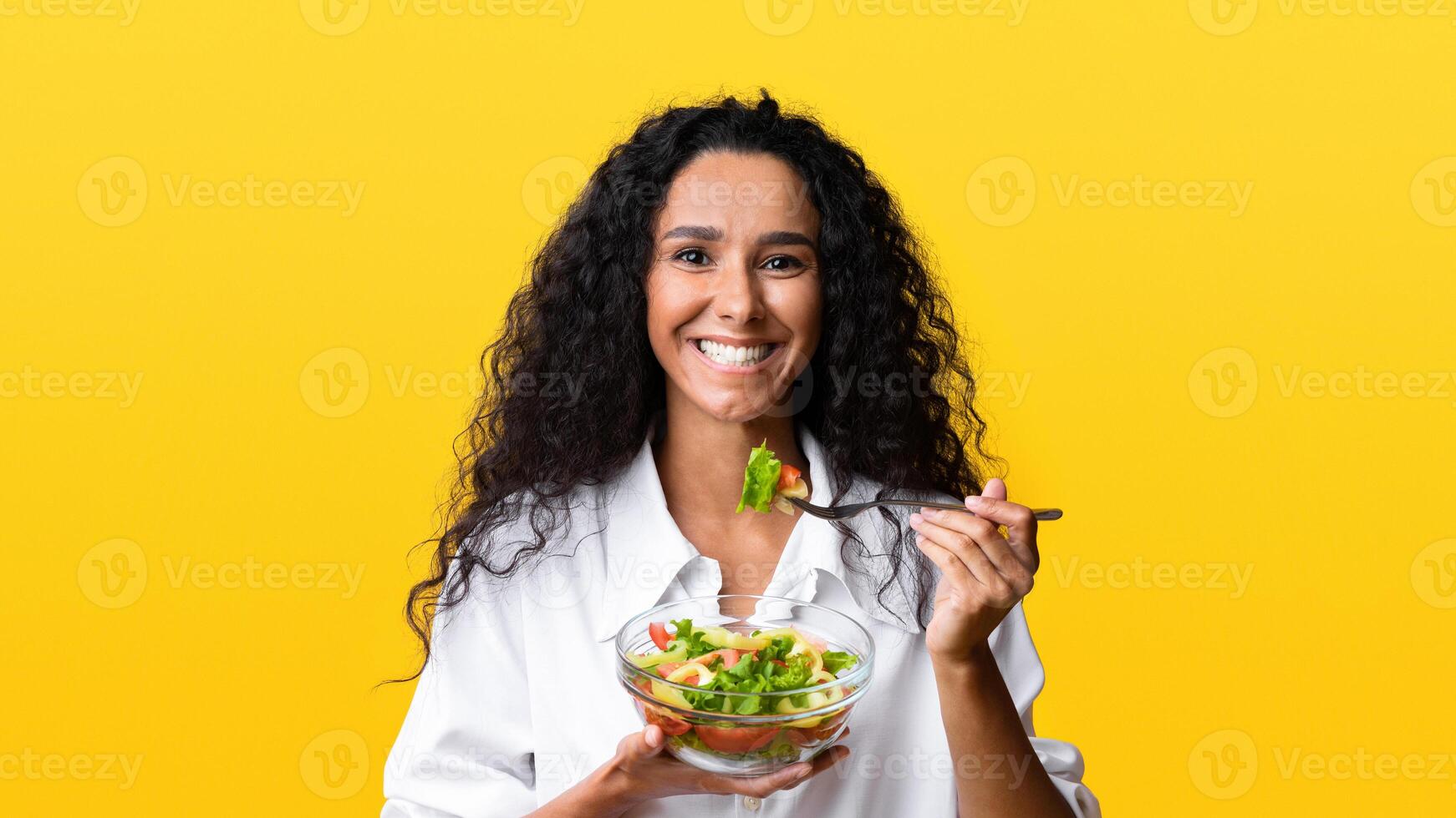 Healthy Eating. Smiling curly woman enjoying fresh vegetable salad over yellow studio background ...