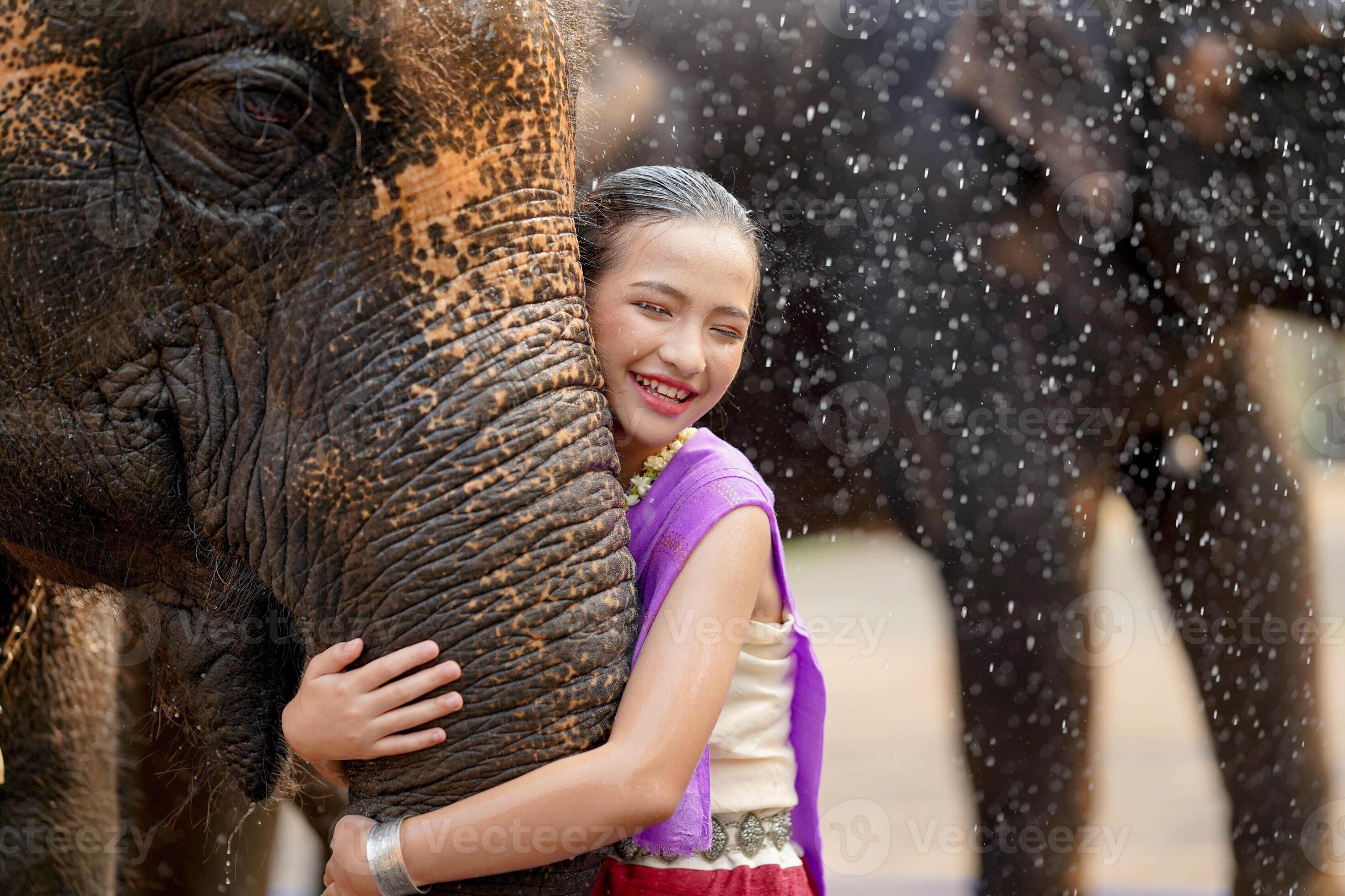 A cheerful Asian girl in the Thai traditional dress hug trunk of Asian elephant in Thailand ...