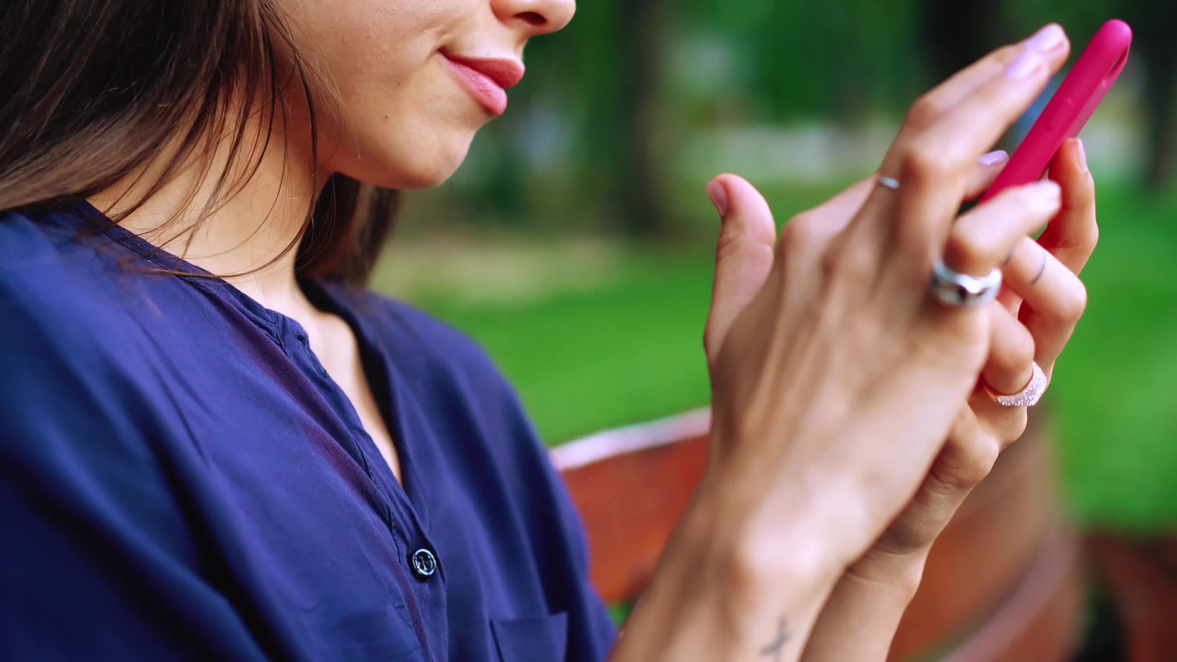 Young brunette woman uses smart phone scrolling and tapping screen on a sitting on a park bench ...