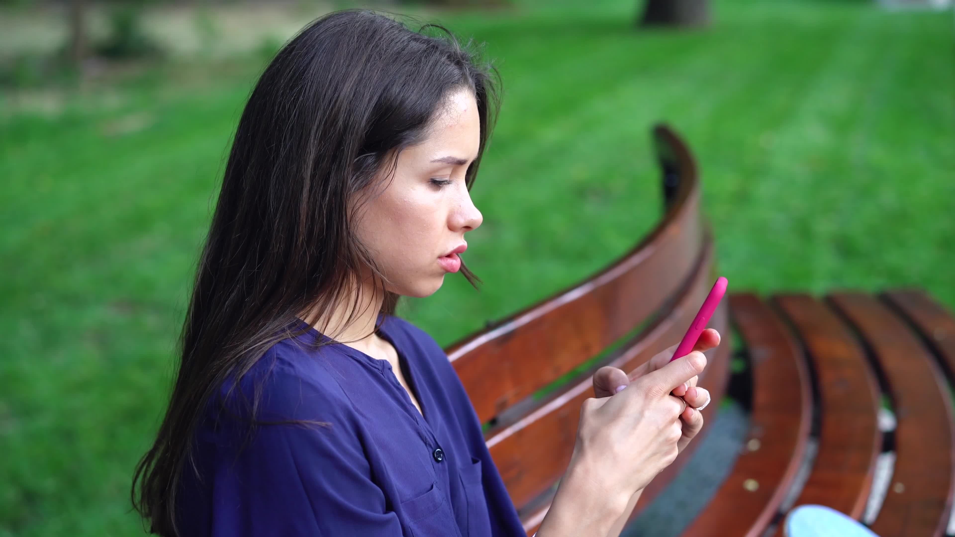 Young brunette woman uses smart phone scrolling and tapping screen on a sitting on a park bench ...