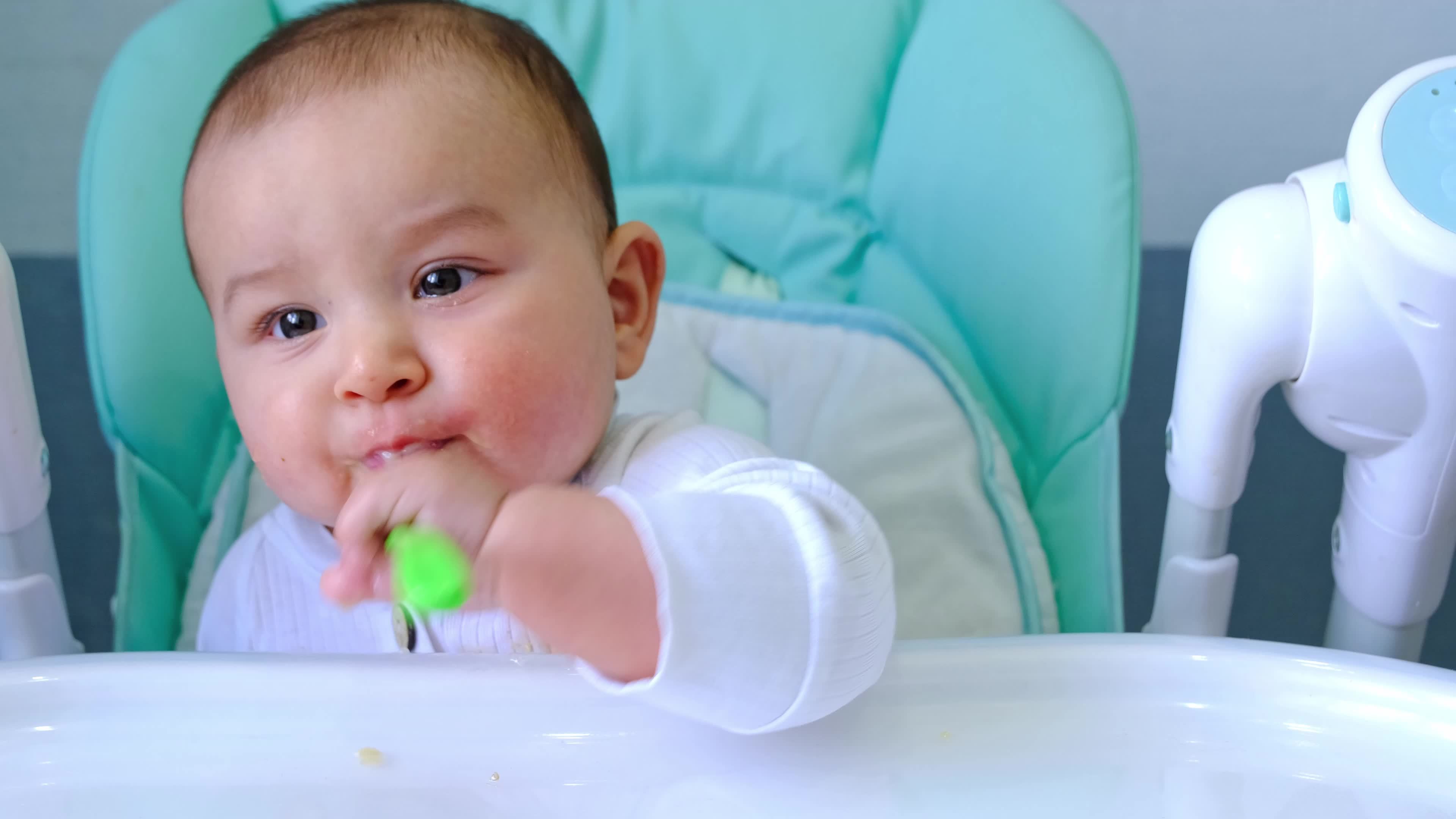 A Hungry Baby Is Gnawing On A Plastic Spoon At The Table On A High a-hungry-baby-is-gnawing-on-a-plastic-spoon-at-the-table-on-a-high