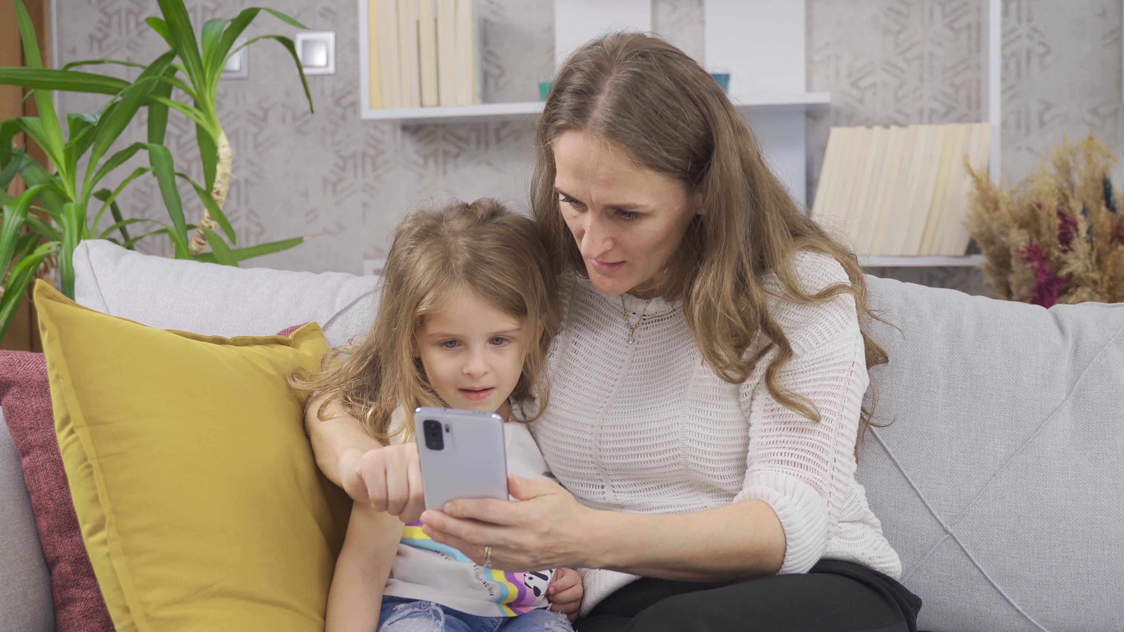 Mother and daughter looking at the phone at home. Mother showing her little daughter something ...