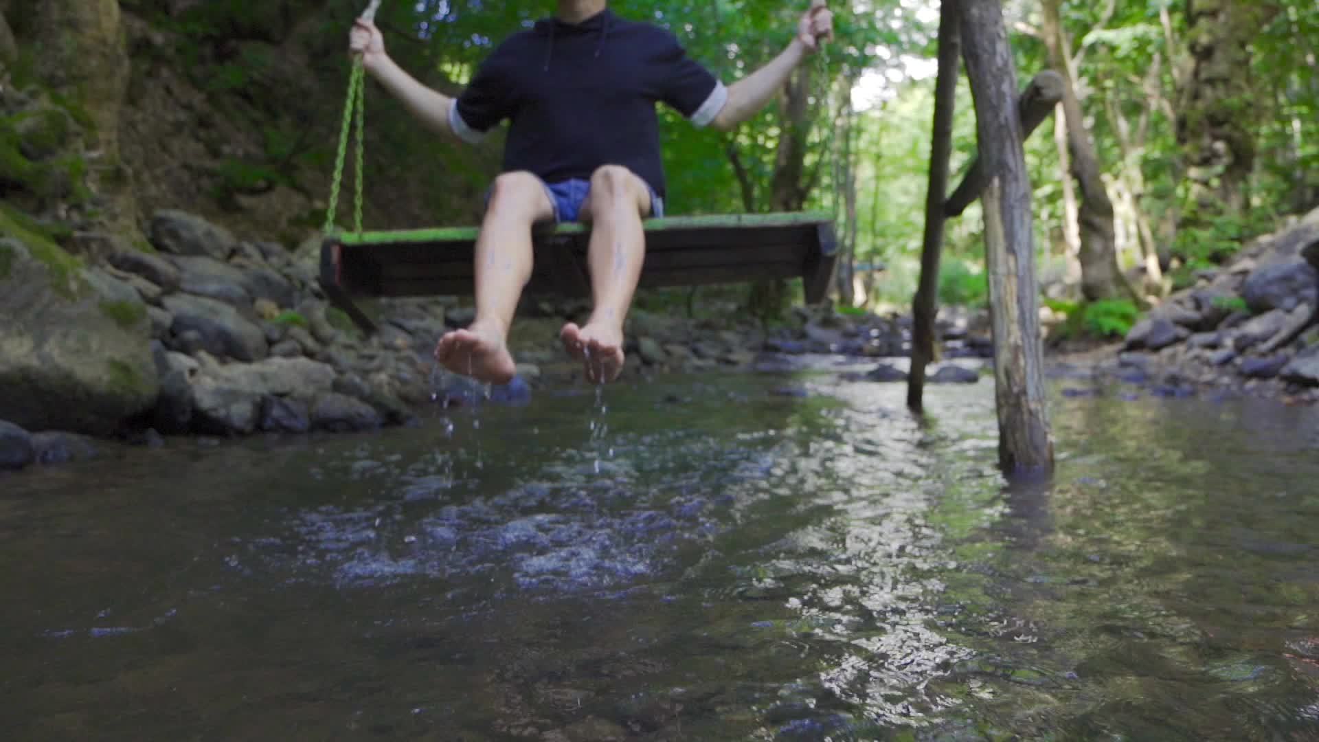 Swinging on a swing over the stream. Slow Motion. Water touches the feet of the young man ...