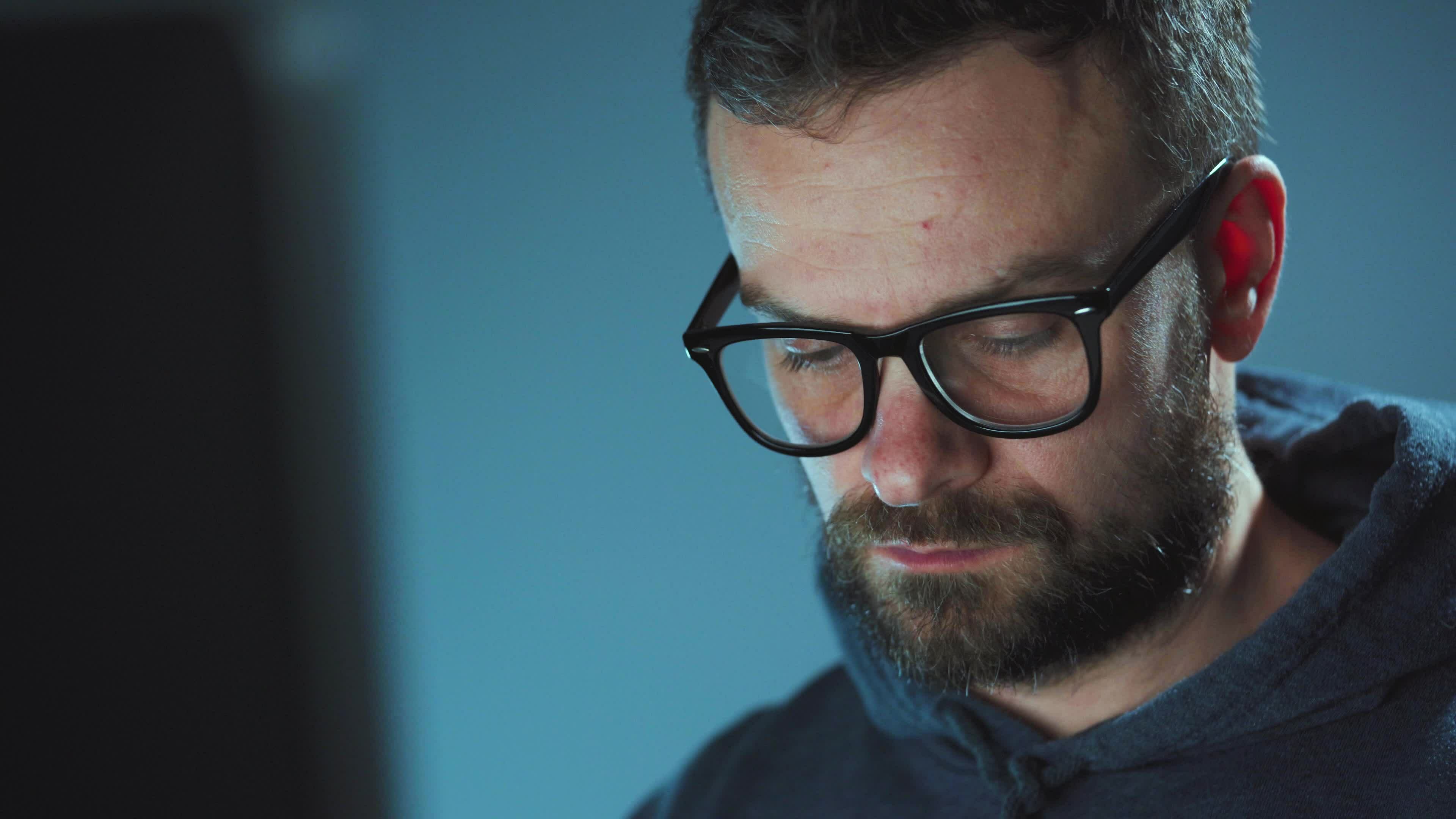 Bearded Male Hacker In Hoodie And Glasses Working On A Computer In A