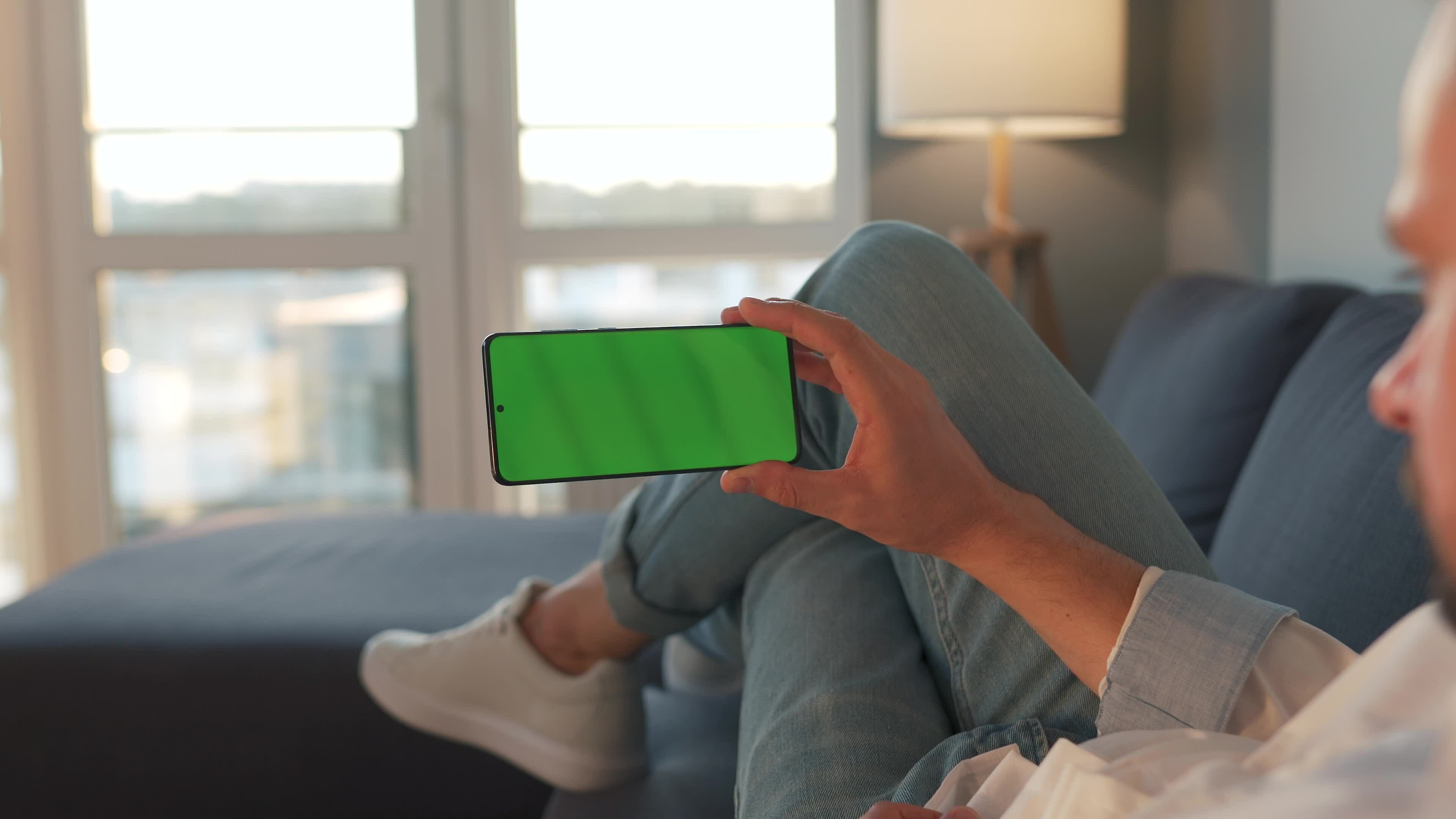 Man at home lying on a sofa and using smartphone with green mock-up screen in vertical mode. Guy ...