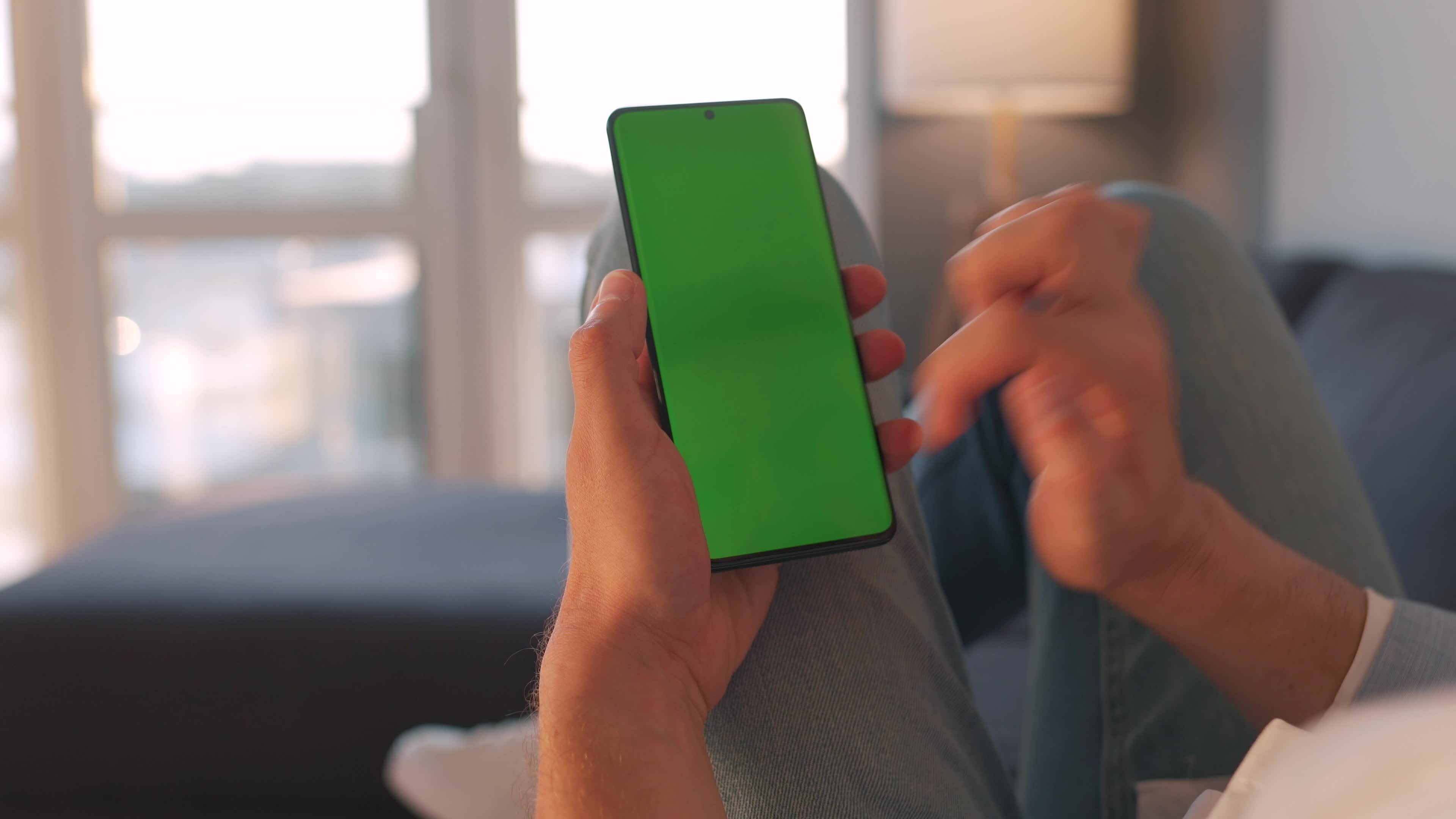 Man at home lying on a sofa and using smartphone with green mock-up screen in vertical mode. Guy ...