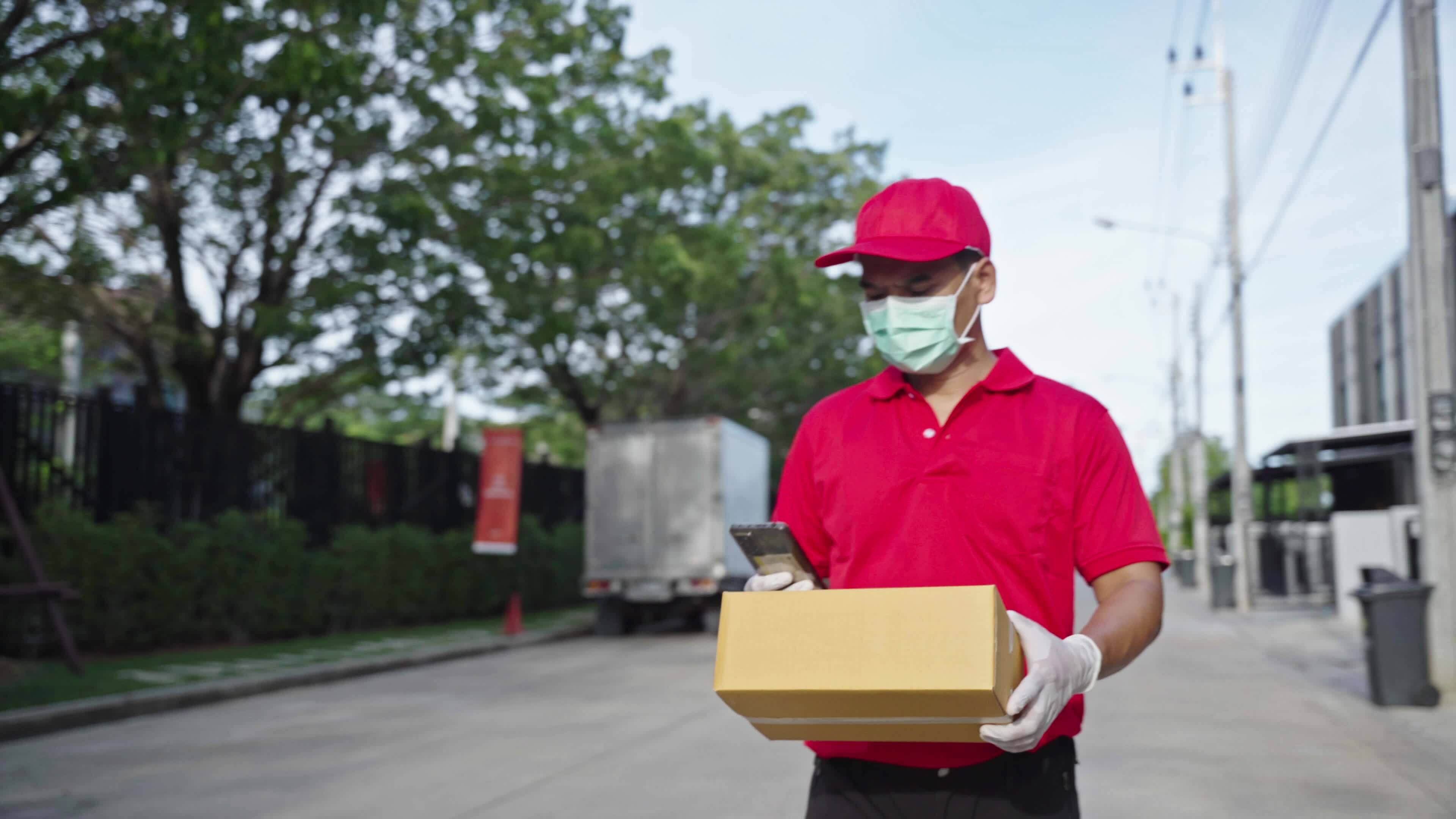 A delivery man wearing a red shirt walks holding a parcel box and using his phone to search for ...