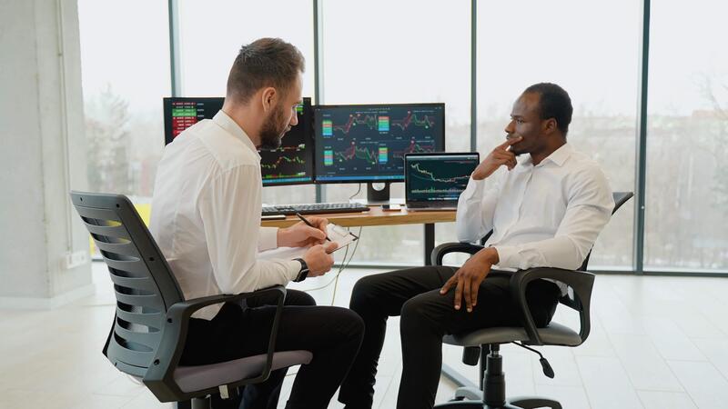 Two crypto traders sitting in front of computers, making professional analysis of candlestick ...