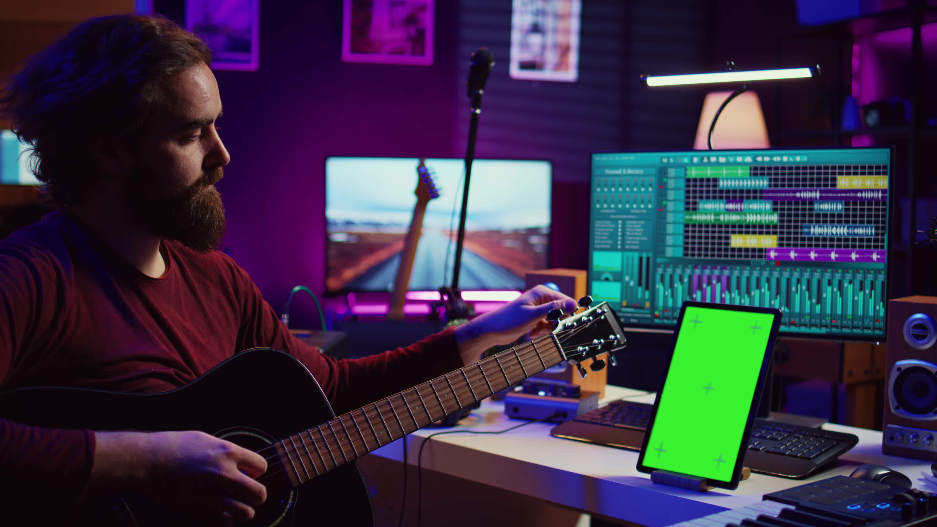 Audio engineer practices tuning his acoustic guitar in home studio, watching a video tutorial ...