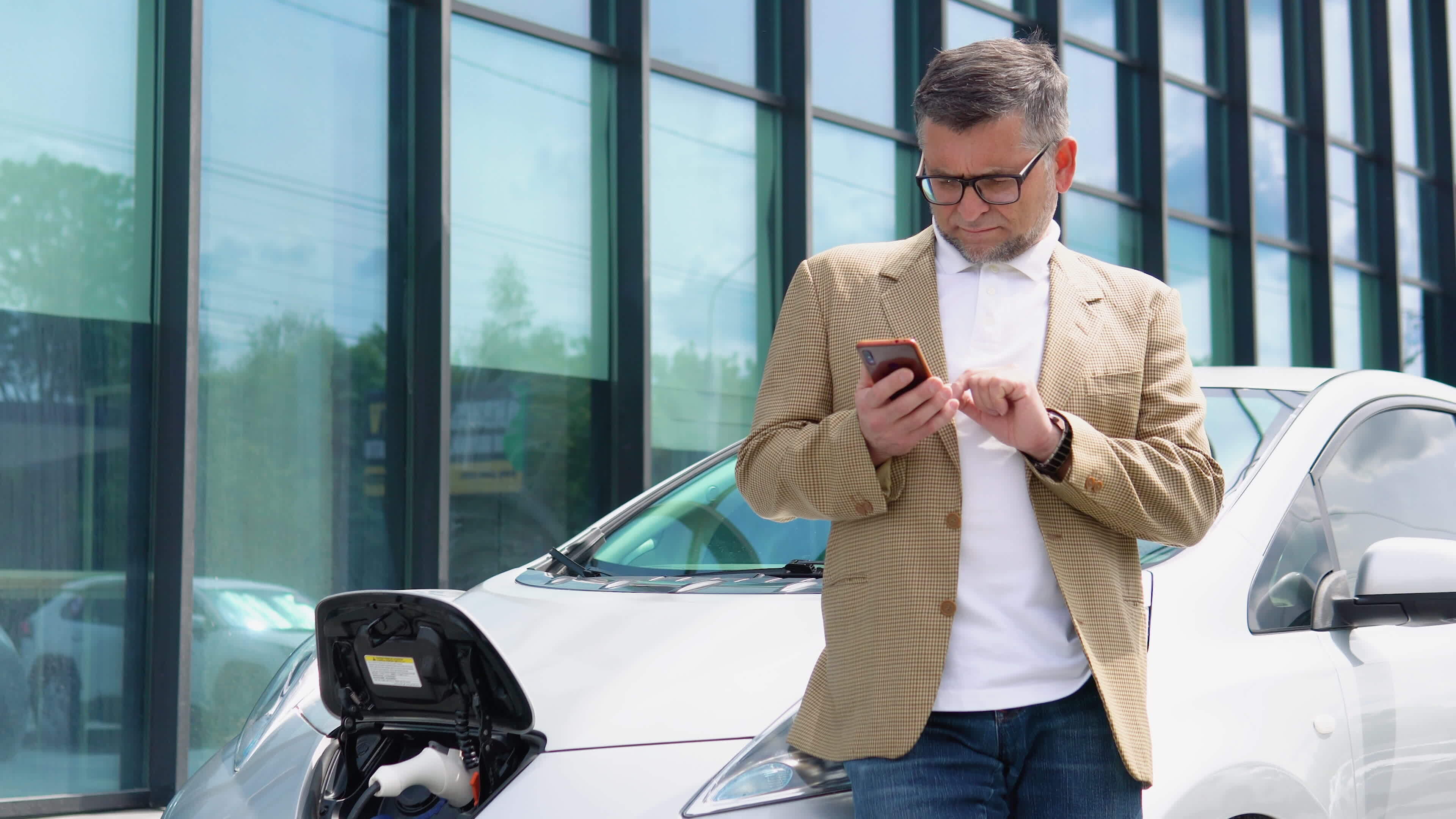 Senior businessman using smart phone and waiting power supply connect to electric vehicles for ...