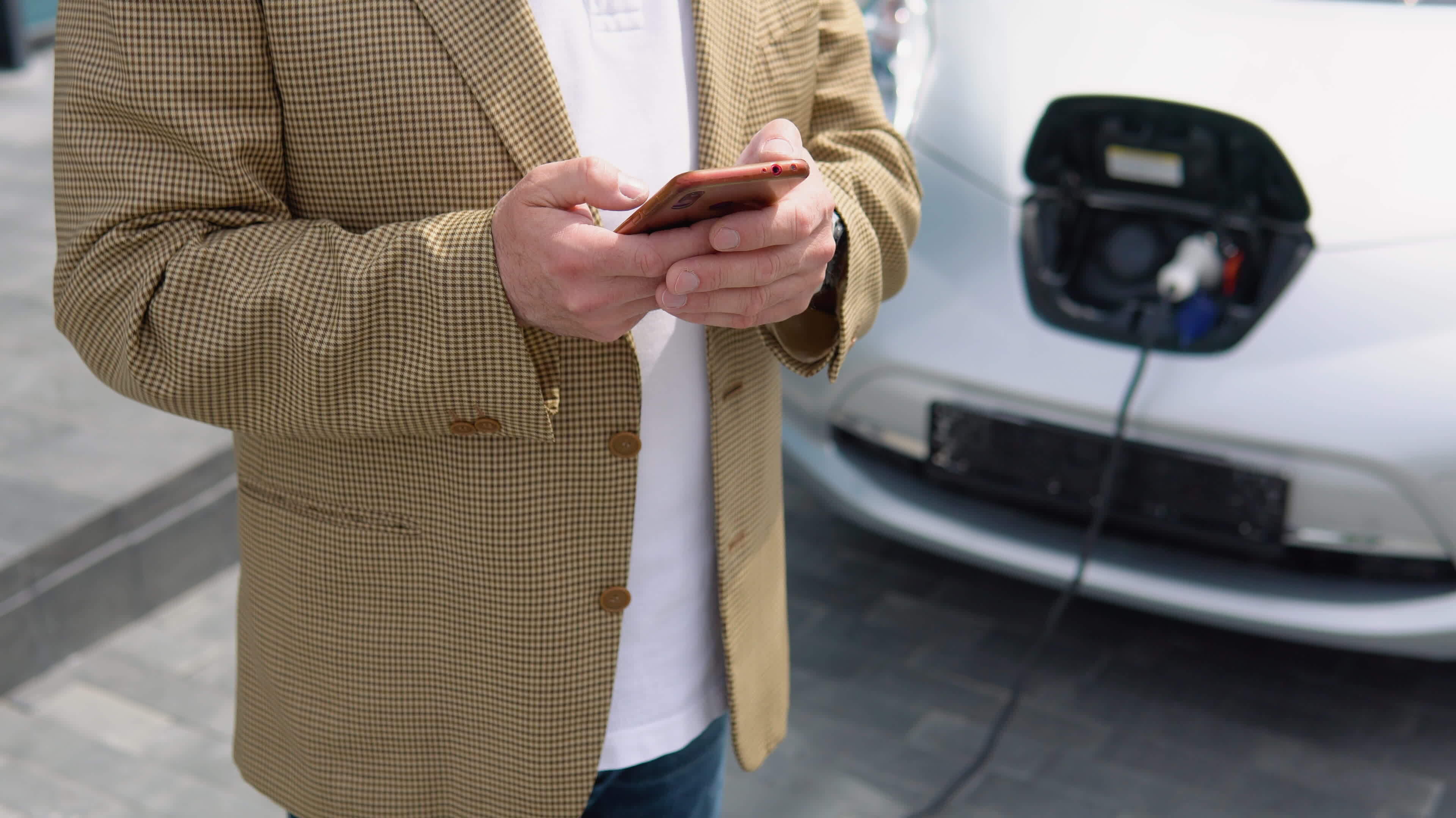Senior businessman using smart phone and waiting power supply connect to electric vehicles for ...