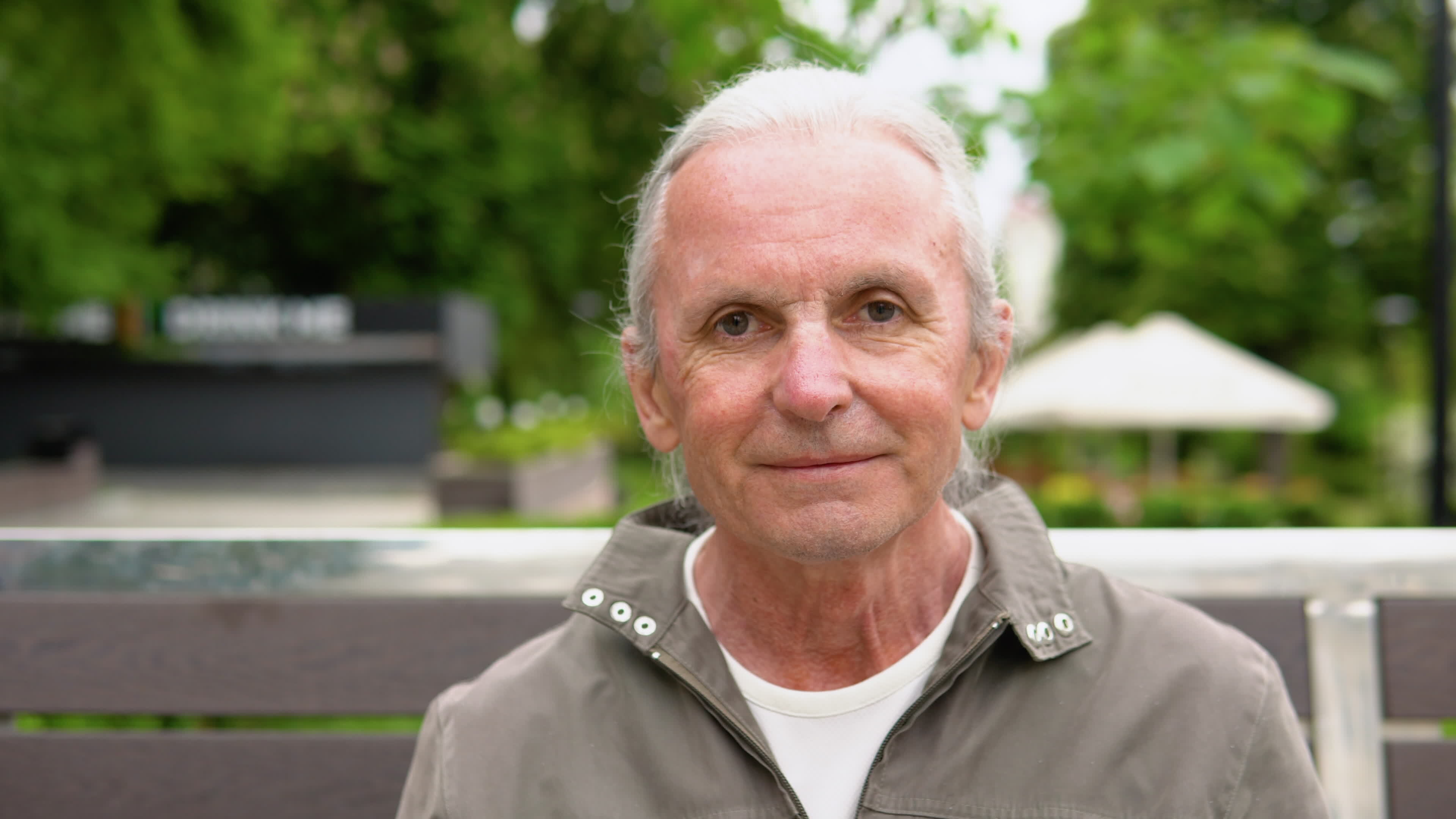 Stylish elderly man with long hair smiling on bench in public city park ...