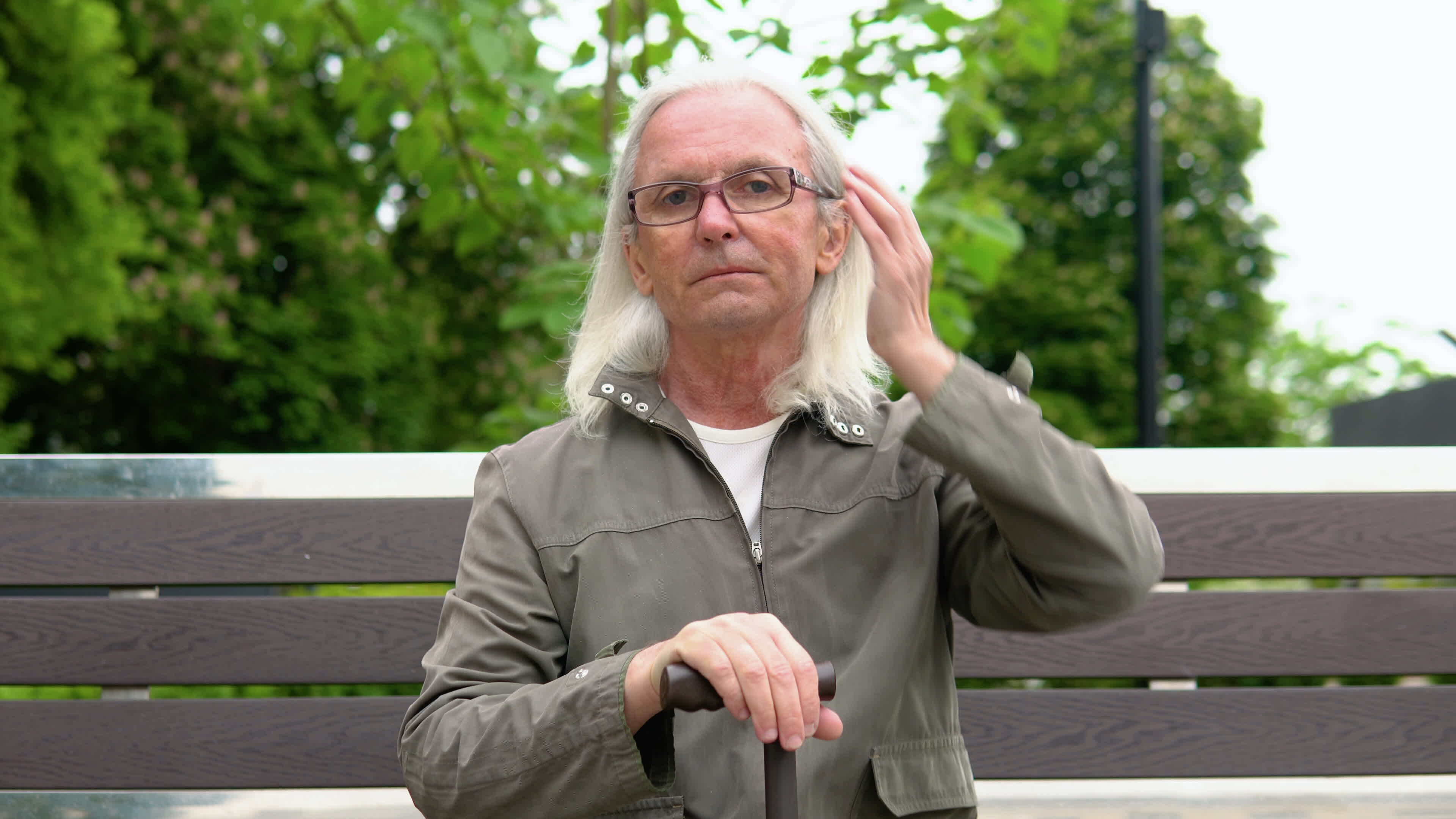 Stylish elderly man with long hair smiling on bench in public city park ...