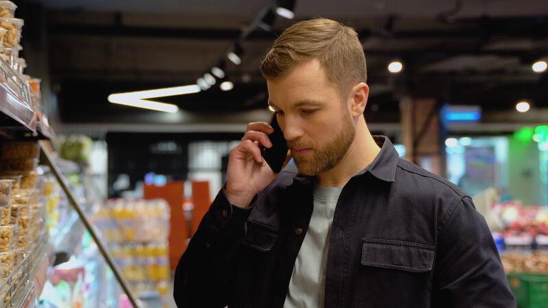 Young caucasian man in a supermarket makes purchases, chooses products and talks on the phone ...