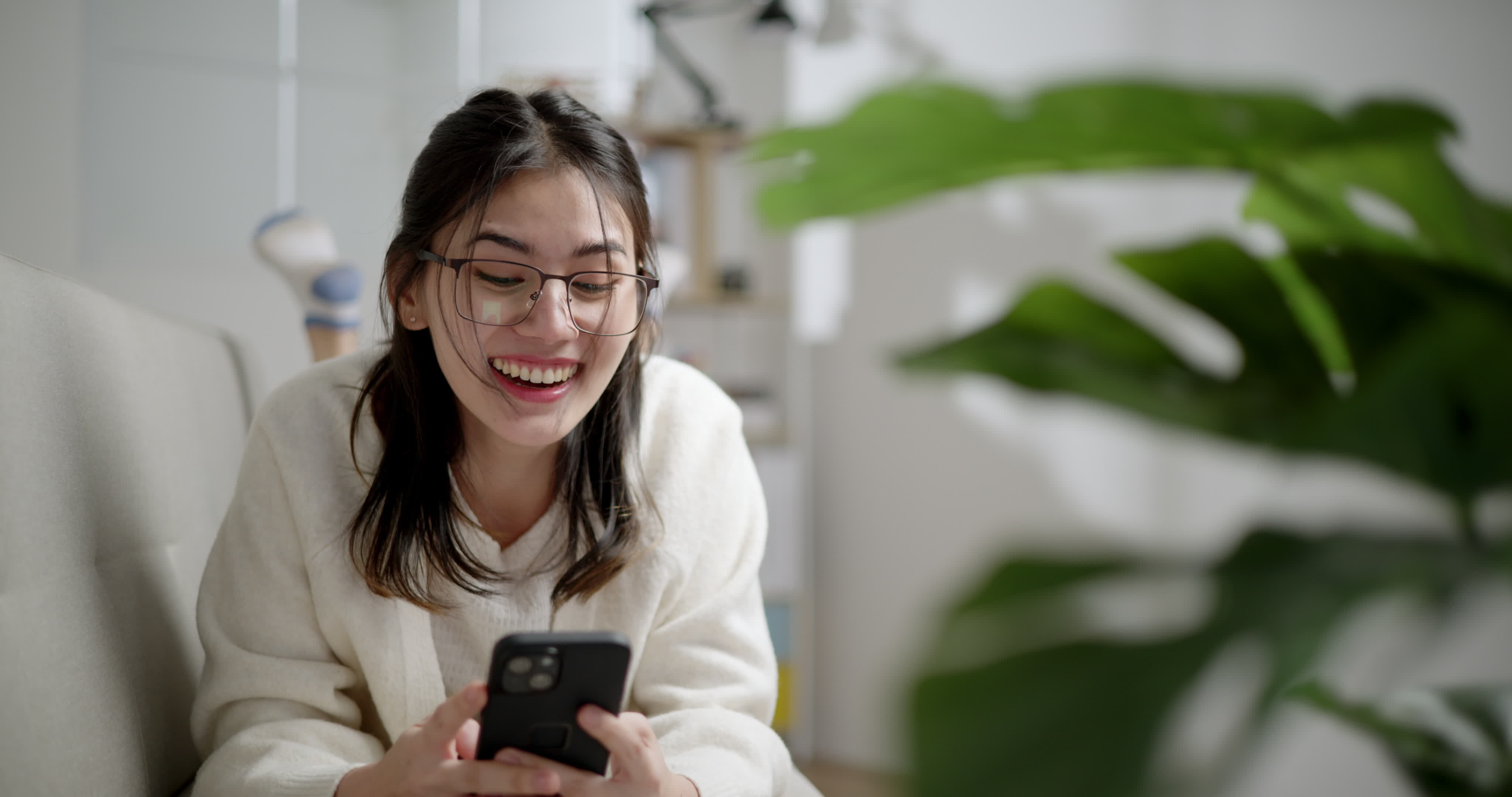 Handheld shot, Young pretty woman in eyeglasses lying on sofa in living room and making online ...