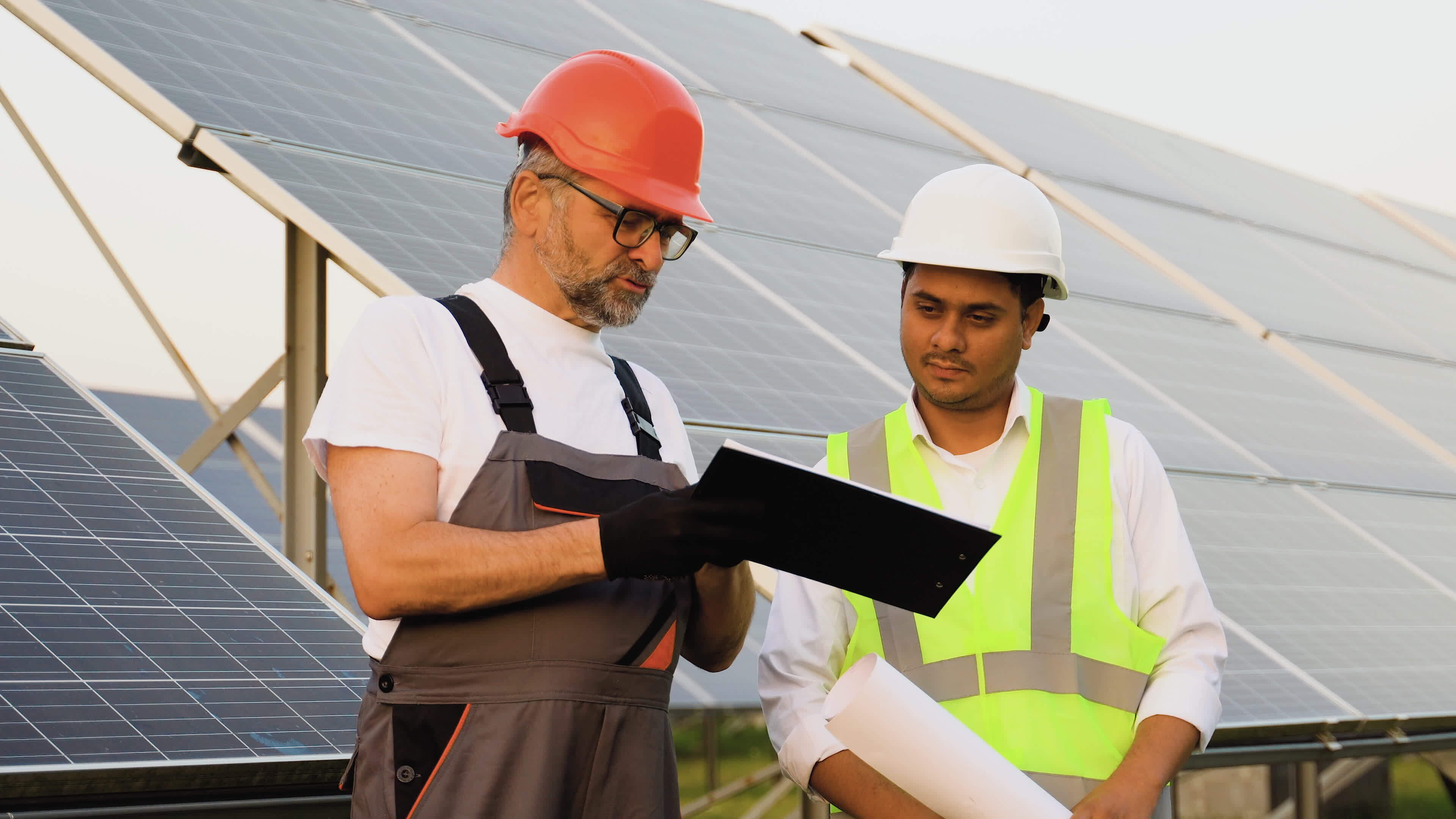 Indian manager and senior worker standing by solar panels with a clipboard, checking the ...