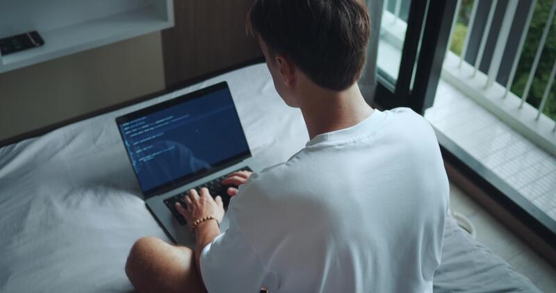 Rear view man freelancer programmer sitting on bed in bedroom and working on laptop computer ...