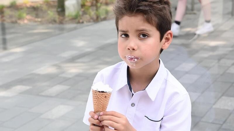 Seven year old boy enjoys delicious ice cream during a walk in the park, licks his dirty lips ...