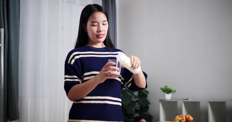 portátil tomada, jovem ásia mulher em pé derramando leite para dentro uma bebendo vidro pronto ...