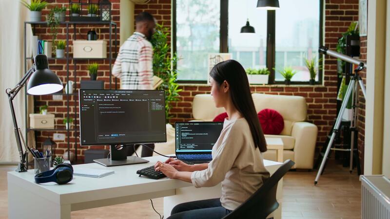 Asian programmer seen typing code for a user interface design on dual monitors, working in a ...