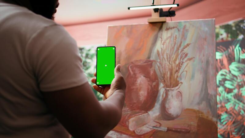 Young artist looking at isolated chroma key display in his painting atelier, using device next ...