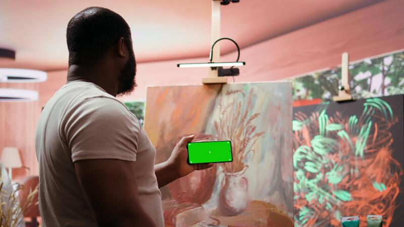 Young artist looking at isolated chroma key display in painting atelier, using device next to ...