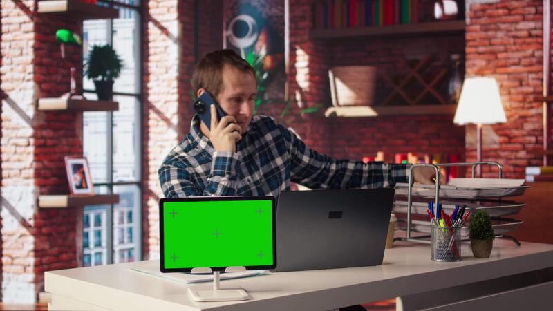 Green screen tablet next to man using laptop, answering phone call while checking emails ...
