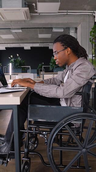 African American freelancer programmer in a wheelchair coding on a laptop at a desk in a modern ...