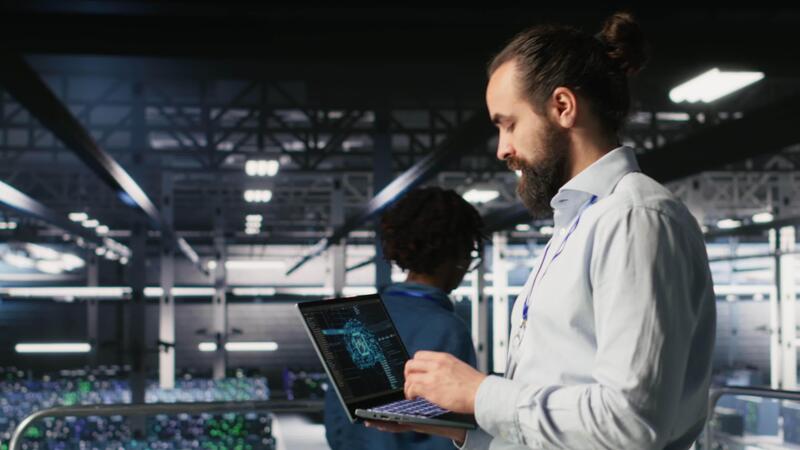 Technician holding laptop walking on server farm platform, doing AI machine learning ...